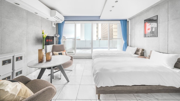 Quarto de hotel com cama, escrivaninha, cadeira, mesa pequena e vista da cidade através de janelas amplas.