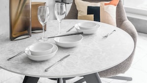 Uma sala de jantar moderna com uma mesa redonda, pratos brancos, talheres, um vaso com flores, uma televisão e um sofá.