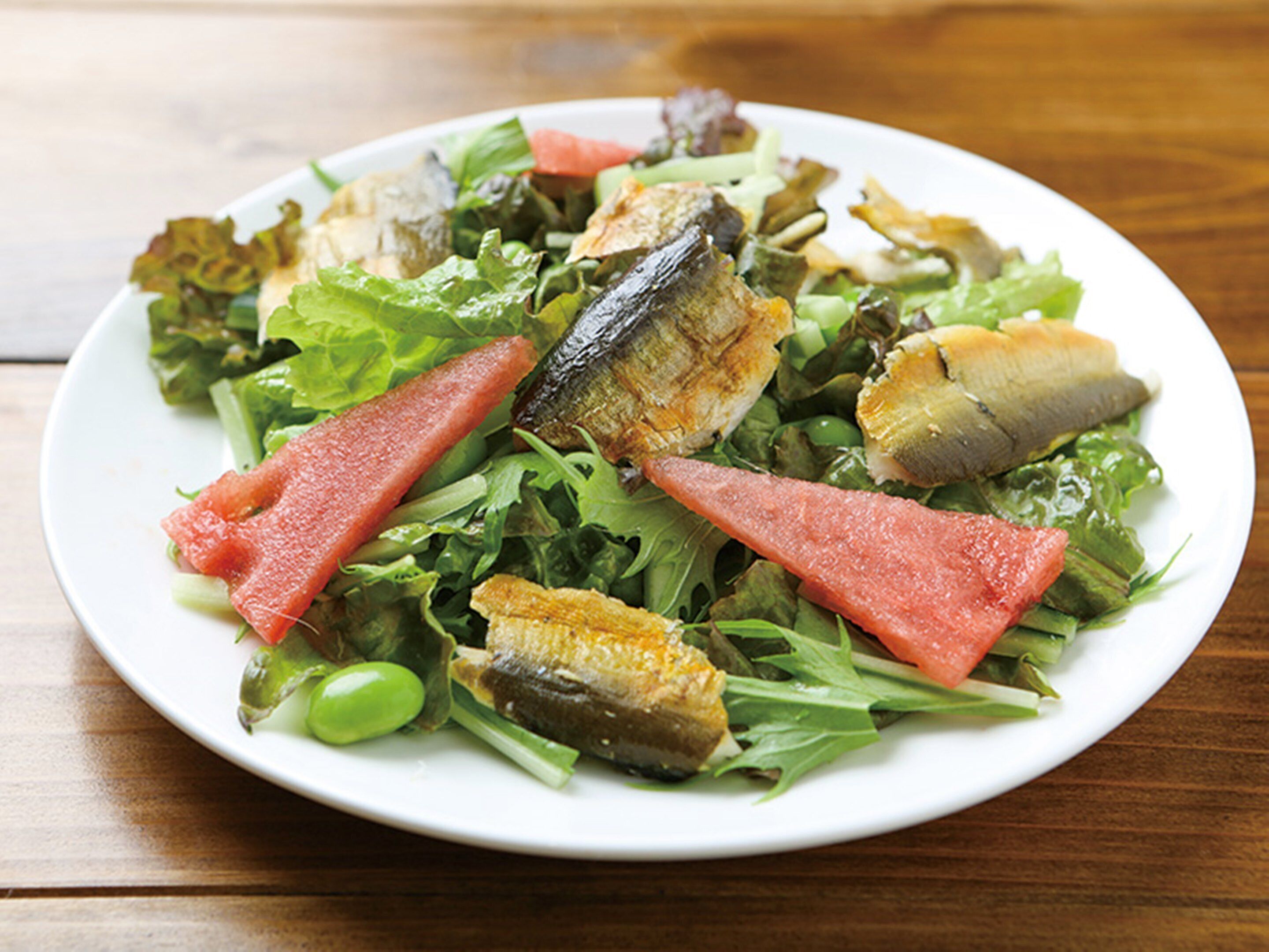 Uma salada com diversas folhas, tomates e peixe sobre uma mesa de madeira.