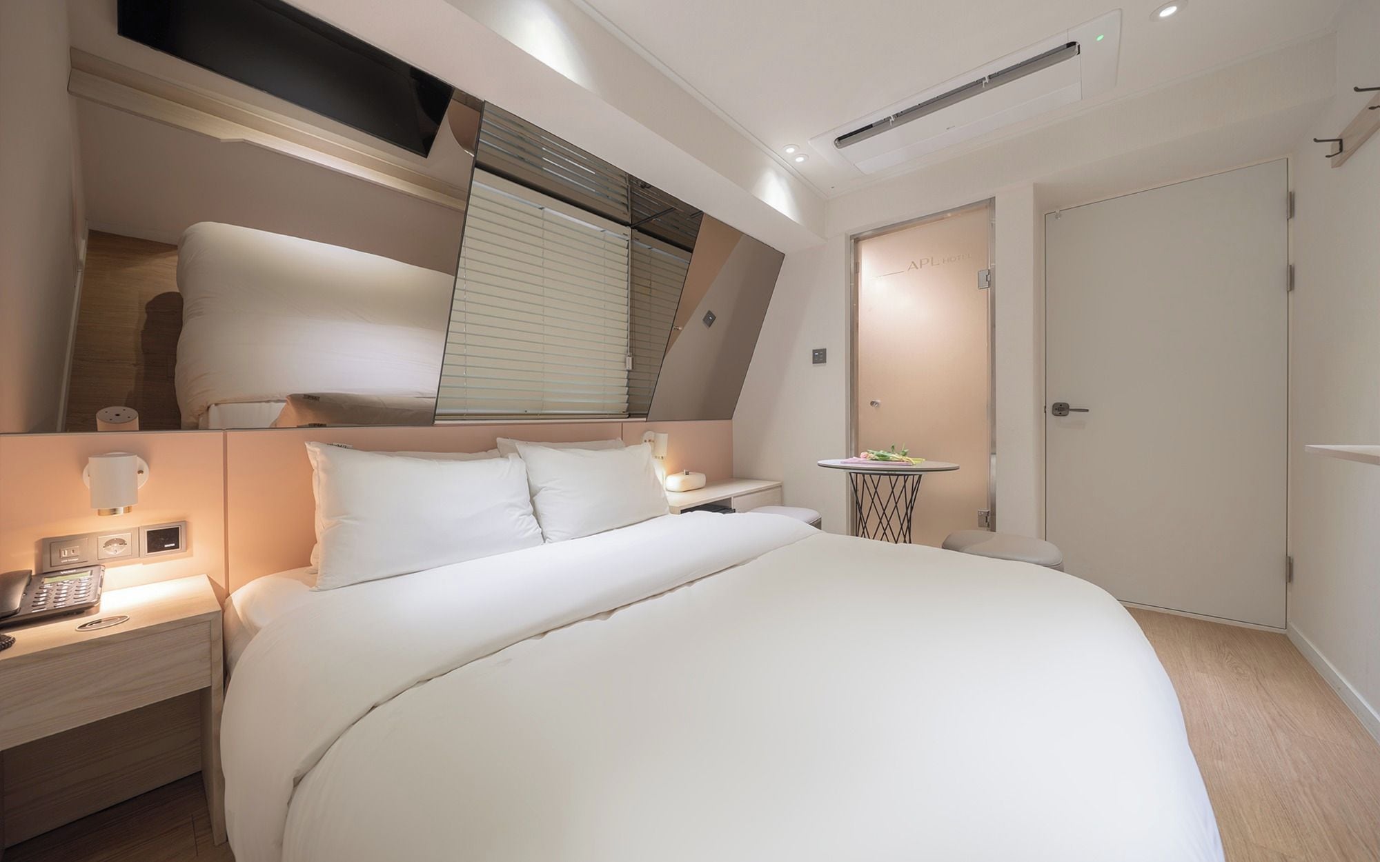 Quarto de hotel moderno com uma cama grande, mesas de cabeceira, uma pequena mesa e uma porta.