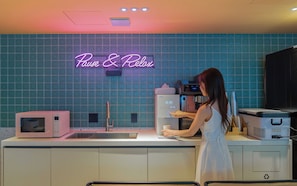 Mulher de vestido branco num balcão de café, com um letreiro de neon que diz 'Pause & Relax'.