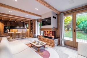 Uma sala de estar moderna com lareira, vigas de madeira, área de cozinha e uma grande porta de vidro deslizante para o exterior.