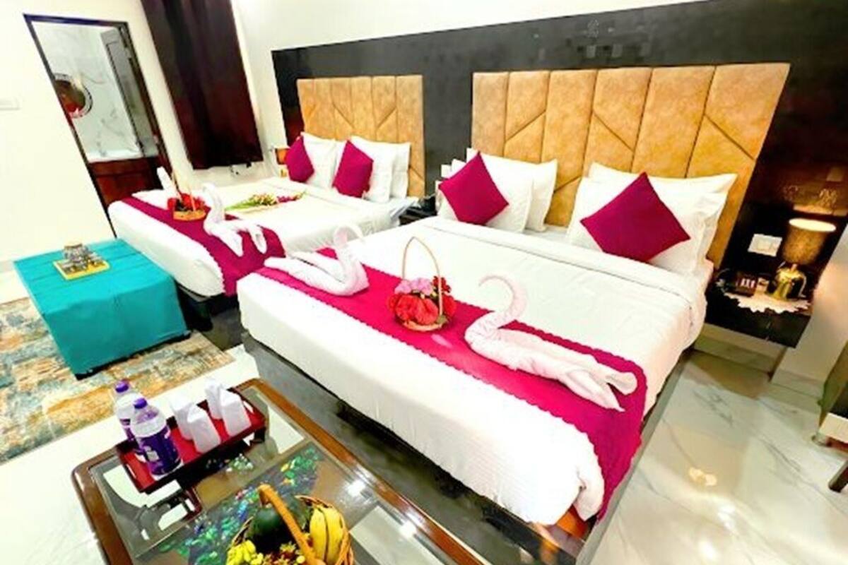 Quarto de hotel com duas camas, uma mesa de centro de vidro com frutas, um banquinho verde-água e um espelho.