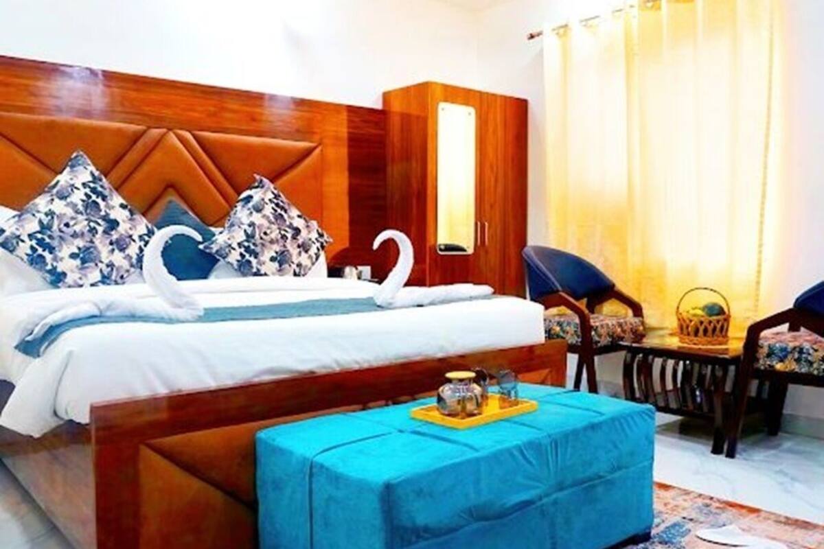 Quarto de hotel com cabeceira de madeira, cama com roupa de cama branca, um banquinho azul e um espaço de jantar com mesa e cadeiras.