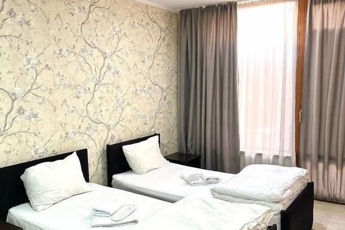 Quarto de hotel com duas camas, uma mesa de cabeceira, uma janela com cortinas e um papel de parede com padrão de árvore.