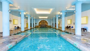 Uma piscina interior ampla com água cristalina, rodeada por colunas e bancos, e uma parede de pedra na extremidade oposta.