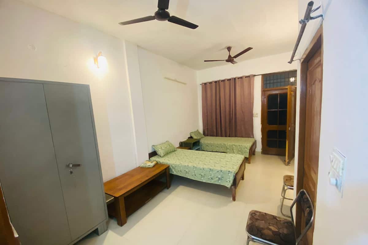 Quarto com duas camas de solteiro, um banco de madeira, uma escrivaninha, uma cadeira, um guarda-roupa e dois ventiladores de teto.