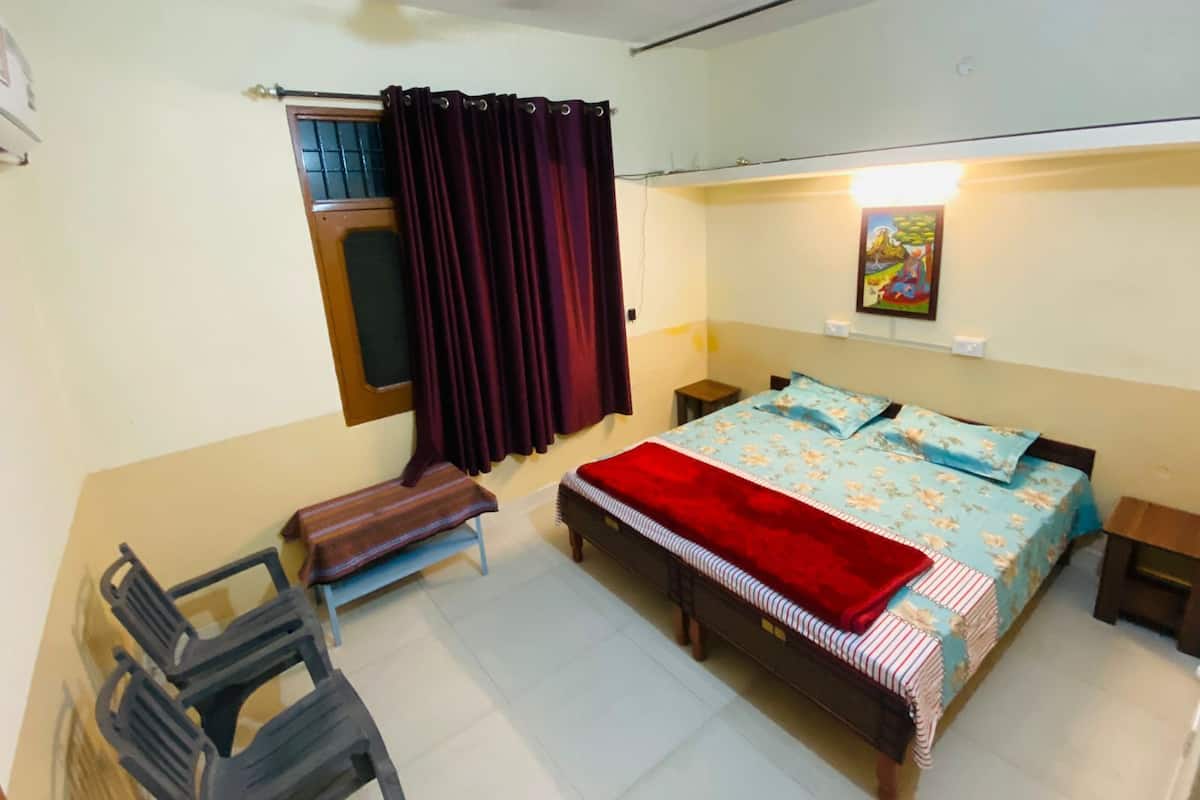 Quarto com cama, um banquinho, janela com cortinas, ventilador de teto e luminária fixada na parede.