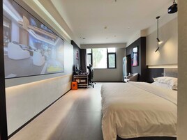 Quarto de hotel moderno com uma cama grande, uma escrivaninha com computador, um projetor e uma janela com vista para o verde.