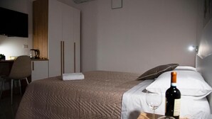 Quarto com cama, mesa de cabeceira com garrafa e copo, cadeira, escrivaninha com cafeteira e televisão fixada na parede.