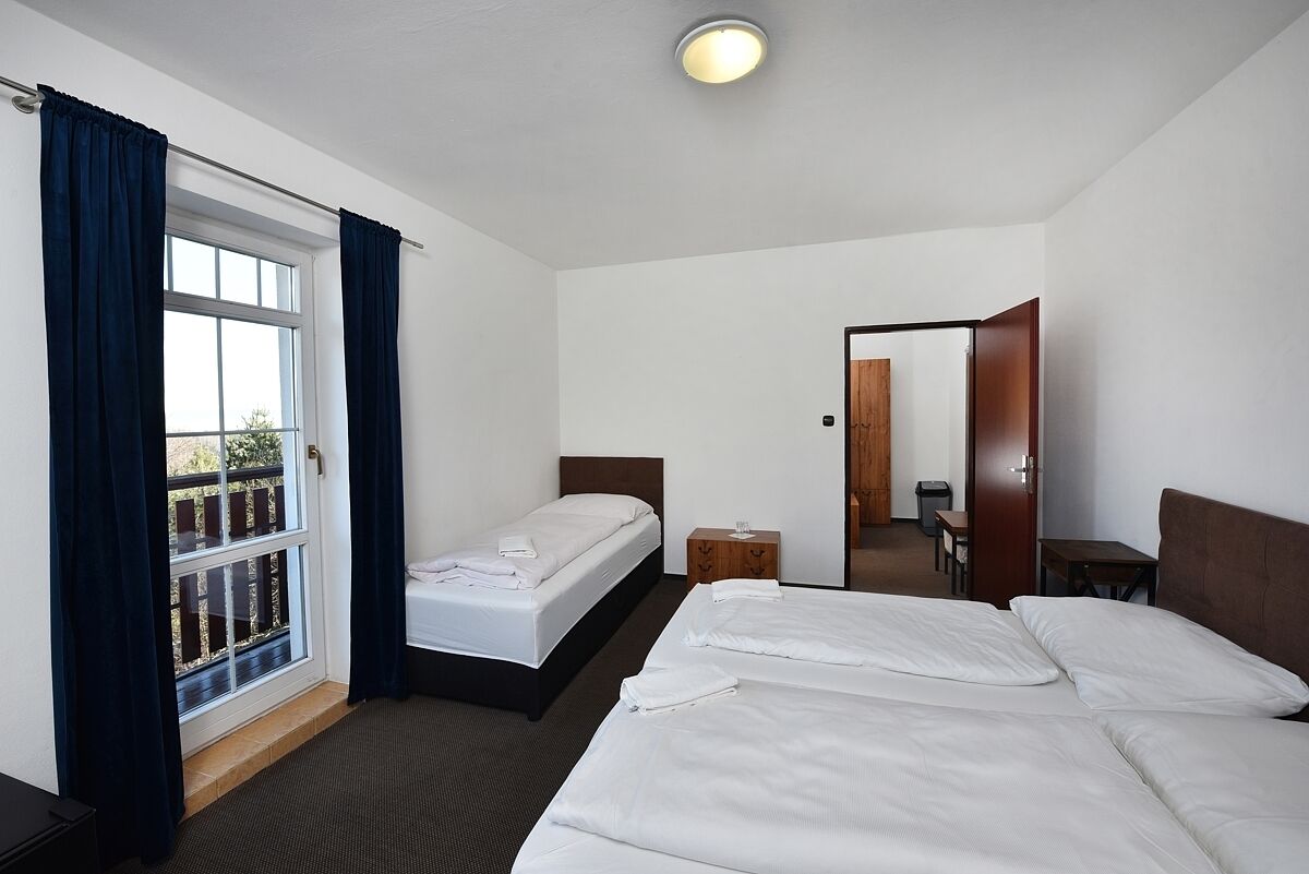 Quarto de hotel com cama, mesa de cabeceira pequena, espelho, janela com cortinas e porta que dá para outro cômodo.