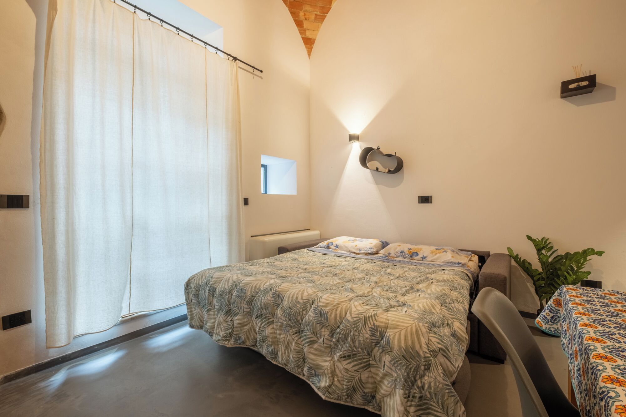 Quarto com cama, janela com cortinas, luminária na parede e uma pequena planta.