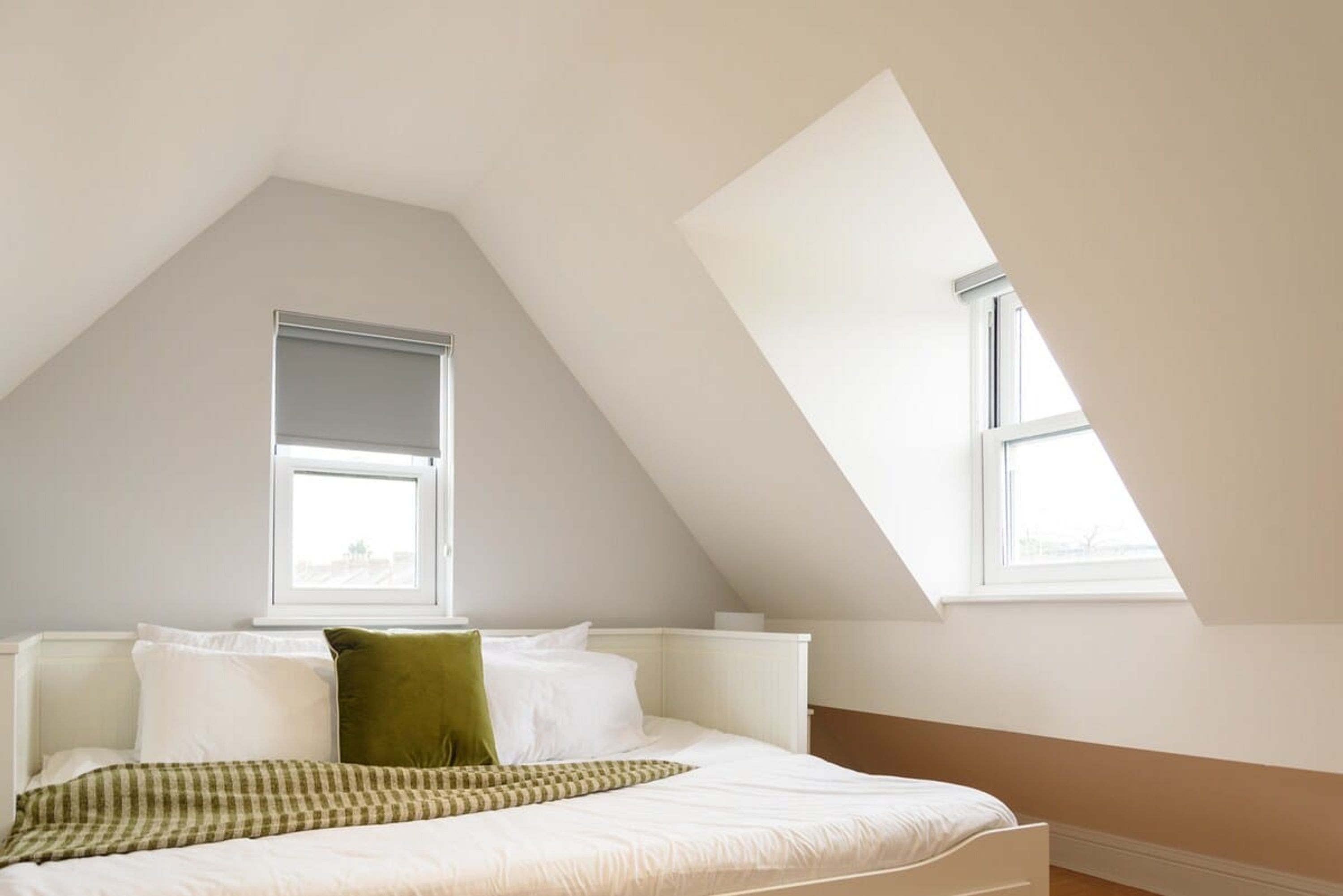Quarto com teto inclinado, cama com roupa de cama branca, travesseiro verde e duas janelas.