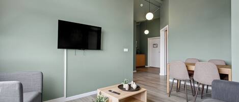 Uma sala de estar moderna com televisão de tela plana, uma mesa de centro com objetos decorativos, um sofá e uma área de jantar com cadeiras e mesa.