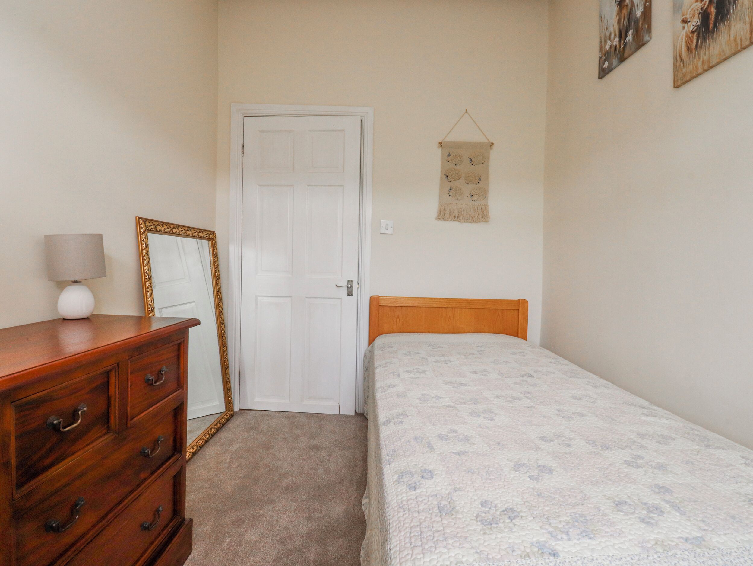 Quarto com cômoda de madeira, espelho, cama com colcha estampada, porta branca e tapeçaria na parede.