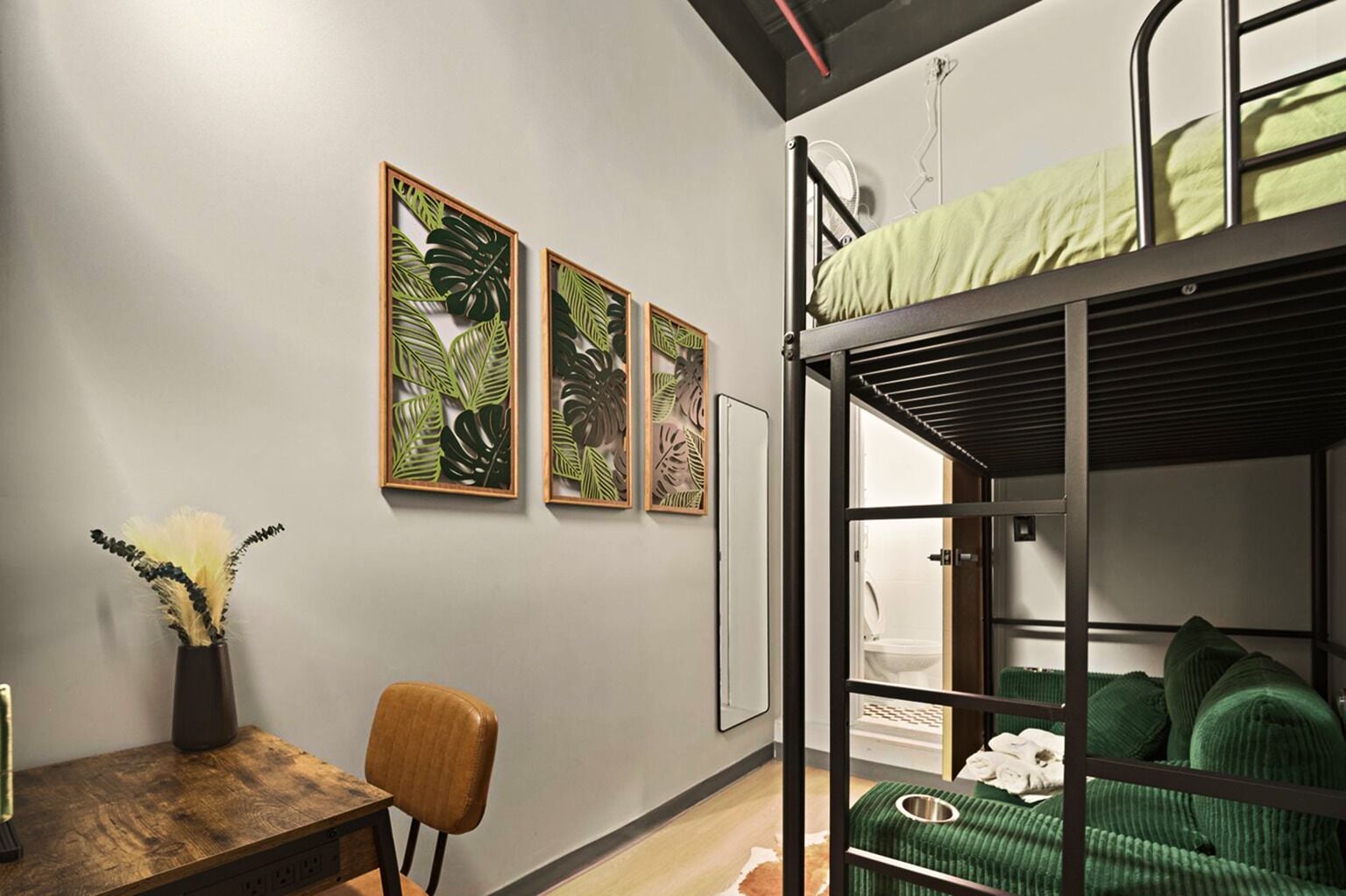 Uma cama de solteiro com roupa de cama verde, uma escrivaninha de madeira com um vaso e três quadros com folhagens tropicais na parede.