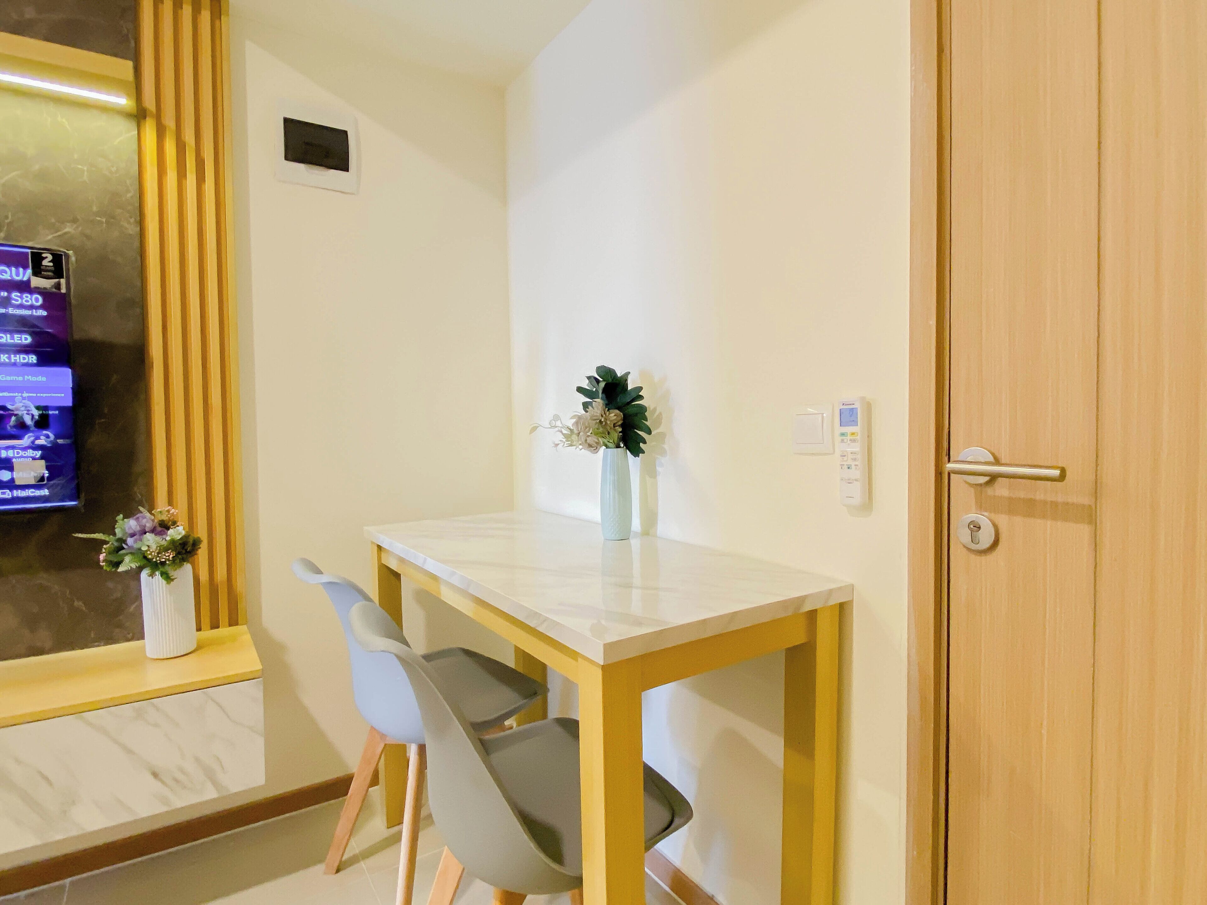 Um quarto pequeno com uma mesa de madeira, duas cadeiras, um vaso com flores, uma televisão na parede e uma porta com tranca.
