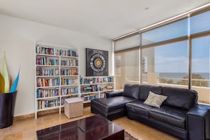 Sala de estar moderna com sofá de couro, estante repleta de livros, um objeto decorativo na parede e uma grande janela com vista.