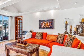 Uma sala de estar com um sofá laranja curvo, uma mesa de centro de madeira e uma grande janela com vista para edifícios.
