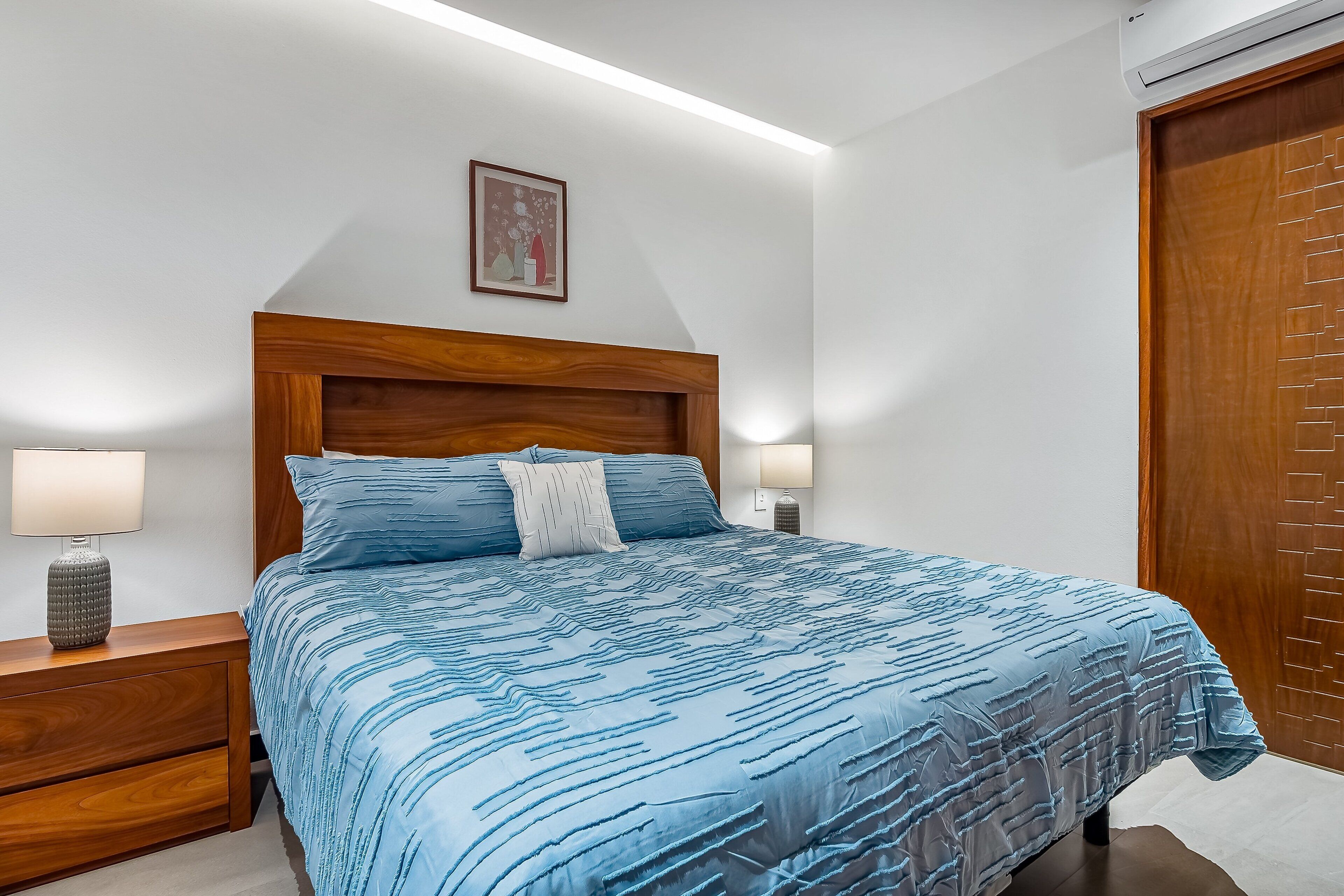 Quarto com cabeceira de madeira, colcha de cama estampada em azul, duas luminárias de apoio e um quadro pendurado na parede.