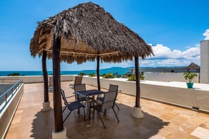 Uma área de estar coberta ao ar livre com um telhado de palha, uma mesa e cadeiras, com vista para a costa.