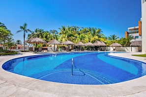 Piscina com escada de metal, cercada por palmeiras e guarda-sóis de palha, com um edifício ao fundo.