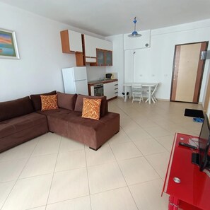 Uma sala de estar com sofá marrom, mesa de jantar com cadeiras, área de cozinha com armários e eletrodomésticos, e uma mesa vermelha com uma televisão.