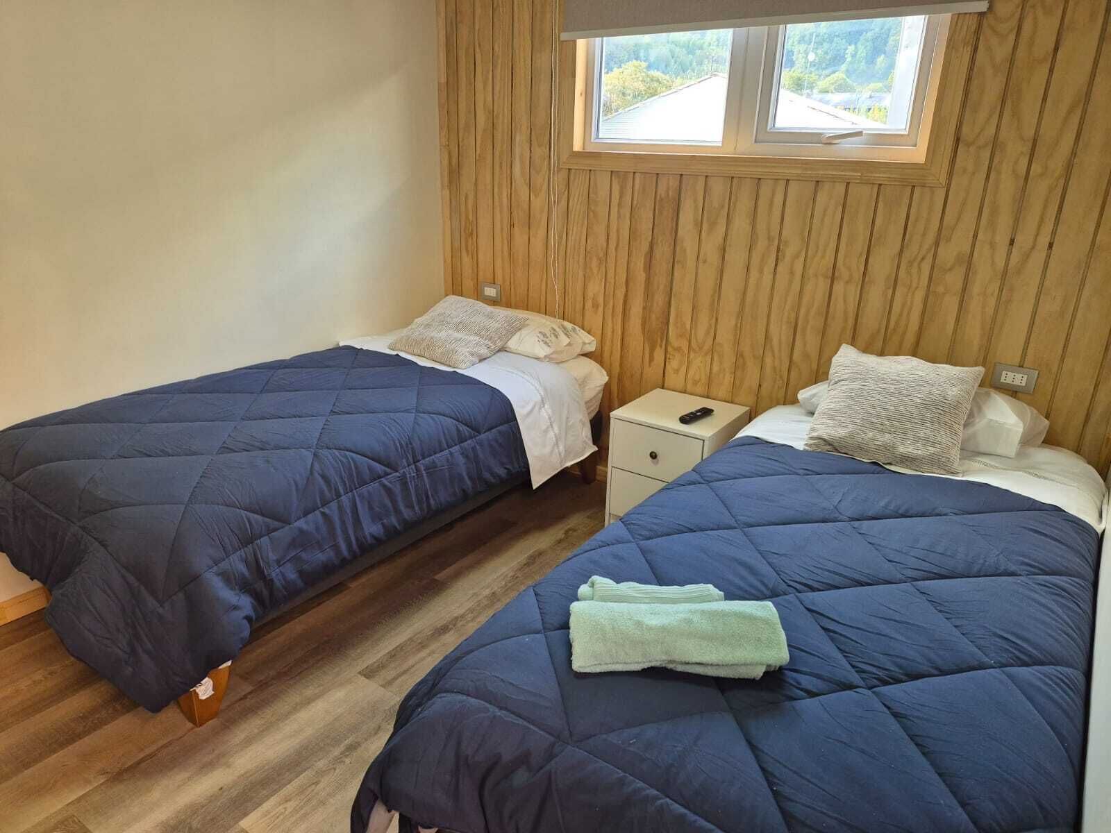 Quarto com duas camas, paredes de madeira, uma janela com vista e uma mesa de cabeceira com controle remoto.