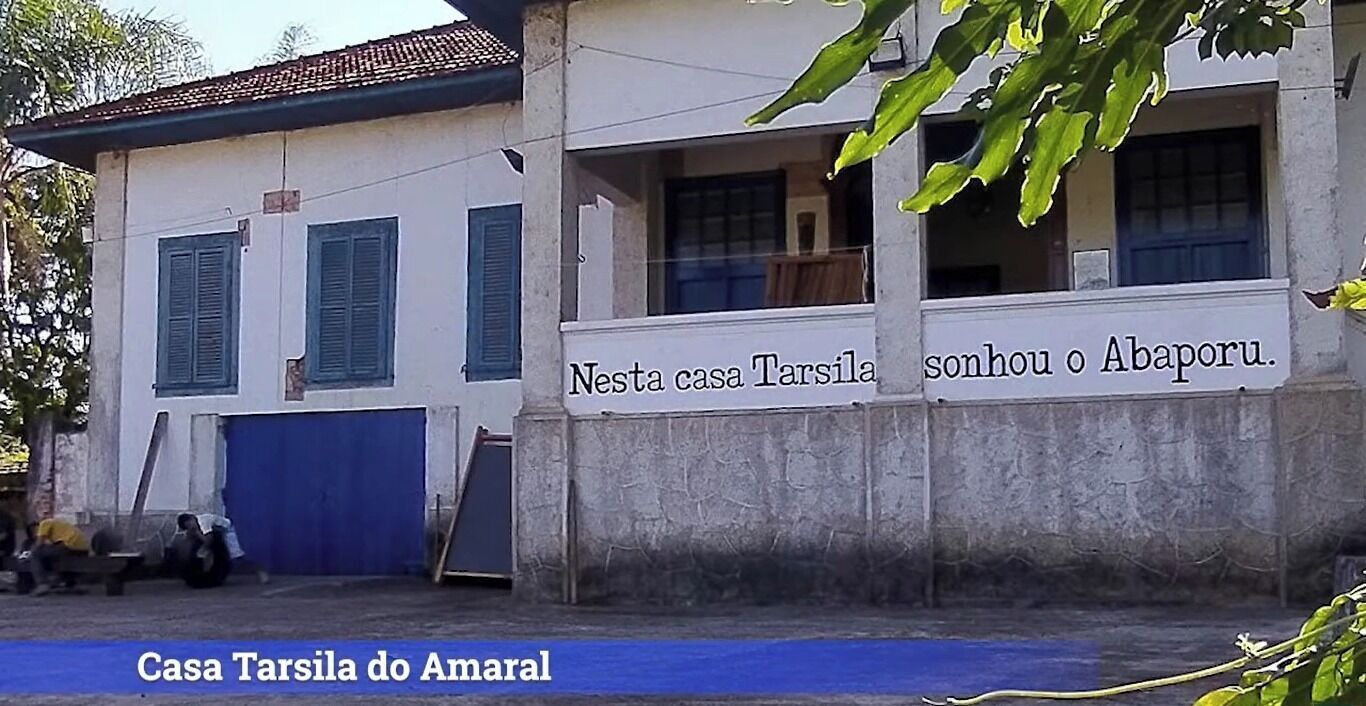 Um edifício de dois andares com uma porta de garagem azul, uma varanda e uma placa indicando que é a Casa Tarsila do Amaral.