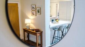 Quarto com mesa de console de madeira, luminária e espelho refletindo uma cozinha com bancada branca e banquetas.