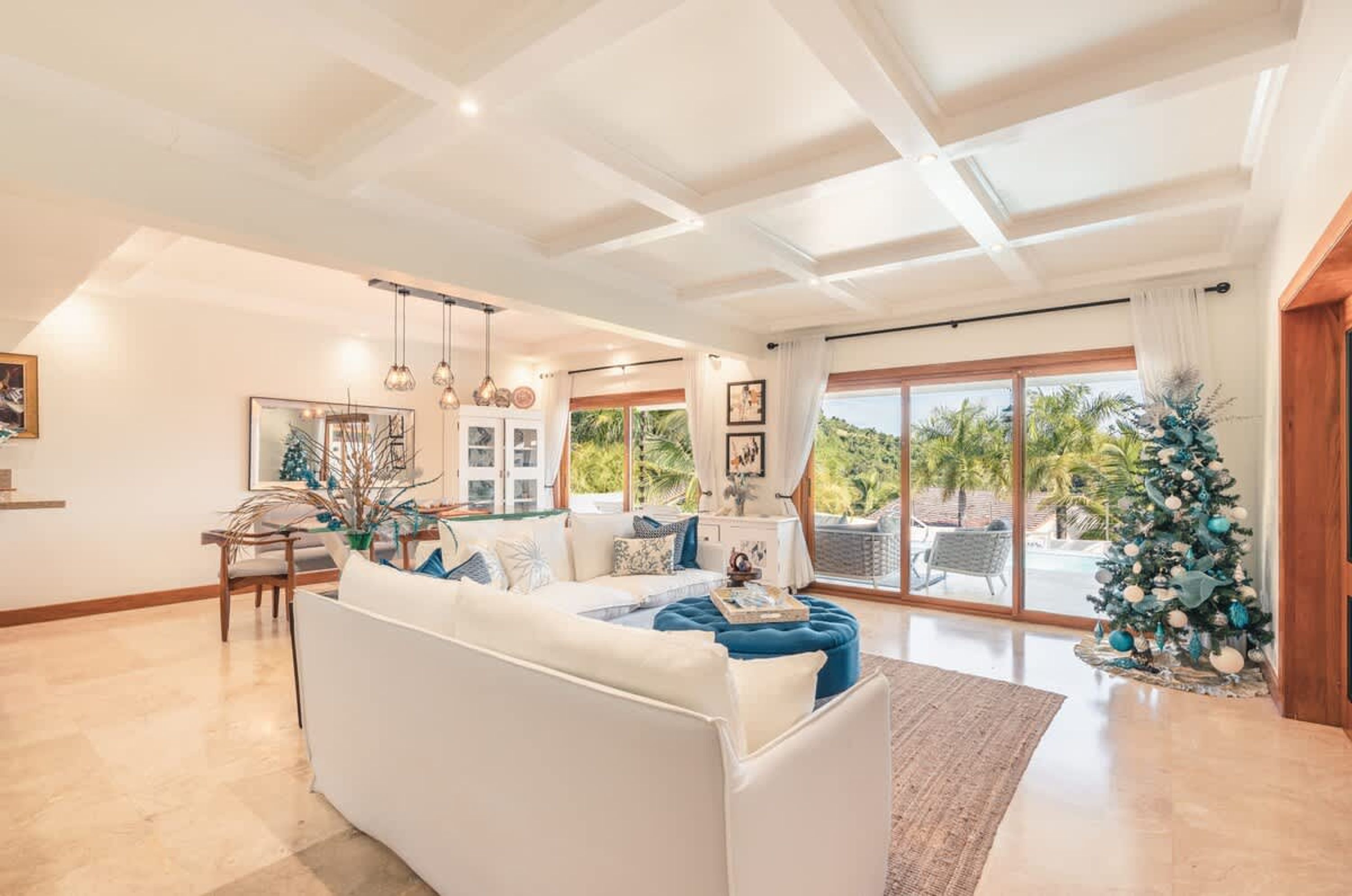 Uma sala de estar espaçosa com um sofá branco, uma área de jantar com mesa e cadeiras, uma janela grande com vista para o verde, e um pinheiro de Natal decorado.