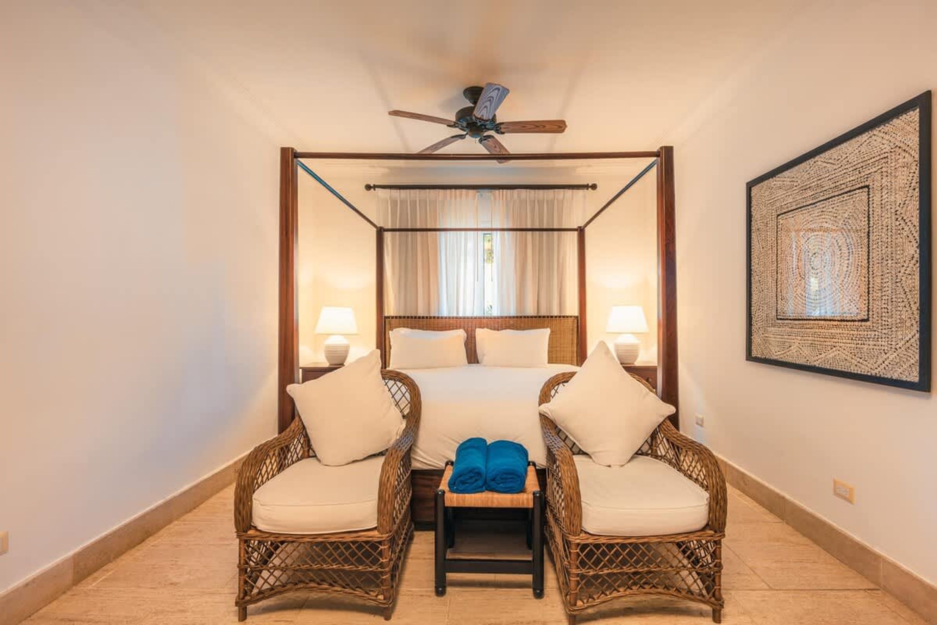 Quarto com cama de dossel, duas cadeiras de vime, uma mesinha com toalhas, um ventilador de teto e uma obra de arte na parede.