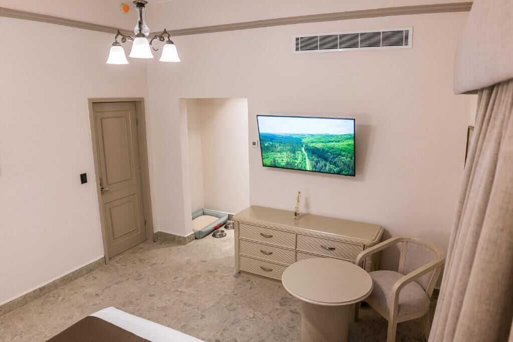 Um quarto com televisão na parede, uma mesinha redonda, um sofá, um armário e uma porta.