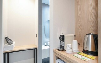 Quarto de hotel com mesa de madeira, máquina de café, chaleira e uma pequena pilha de xícaras brancas.