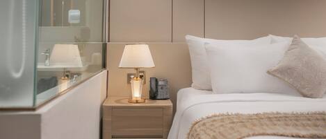 Quarto de hotel com cama, luminária de cabeceira, mesa de apoio e um espelho refletindo o ambiente.