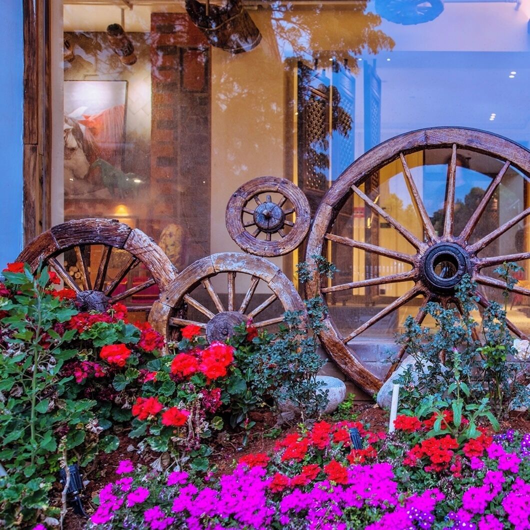 Um jardim com grandes rodas de madeira, flores vermelhas e cor-de-rosa, e a entrada de um edifício.