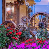 Um jardim com grandes rodas de madeira, flores vermelhas e cor-de-rosa, e a entrada de um edifício.