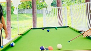 Uma mesa de bilhar com bolas, uma pessoa segurando um taco e vista para um jardim lá fora.