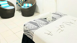 Quarto com uma cama, duas cadeiras de vime com almofadas, uma televisão na parede e uma porta.