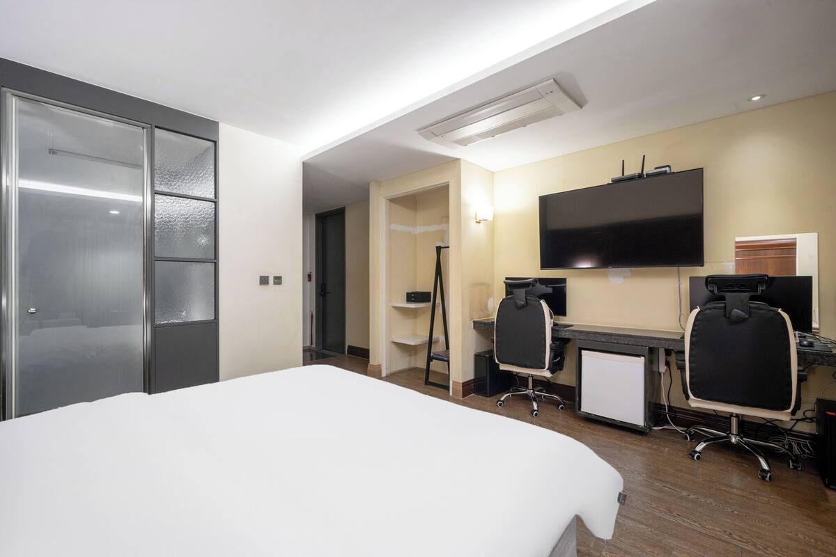 Quarto de hotel com uma cama, uma escrivaninha com duas cadeiras, uma televisão na parede e um guarda-roupa.