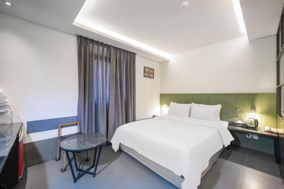Quarto de hotel com cama, escrivaninha, cadeira, mesa de cabeceira e luminária.