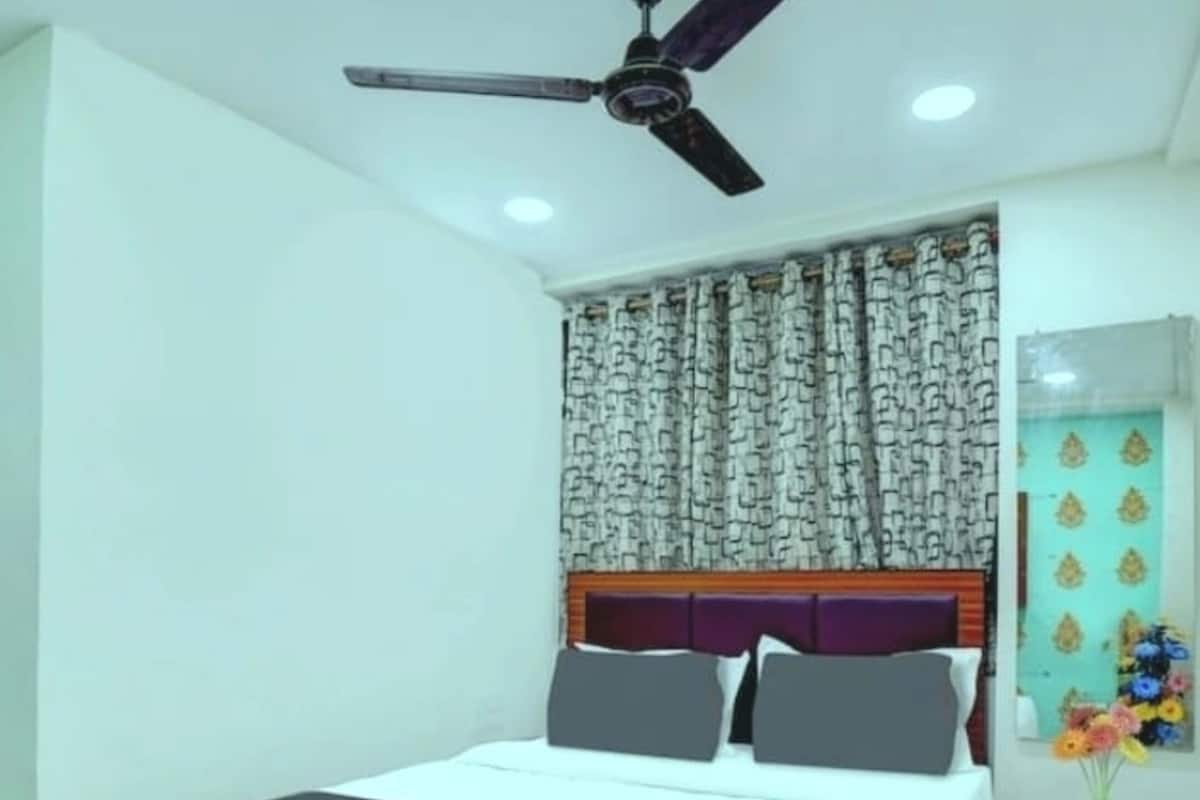 Um quarto com uma cama, um ventilador de teto, uma janela com cortinas estampadas, uma mesa de cabeceira com um vaso de flores e um espelho na parede.