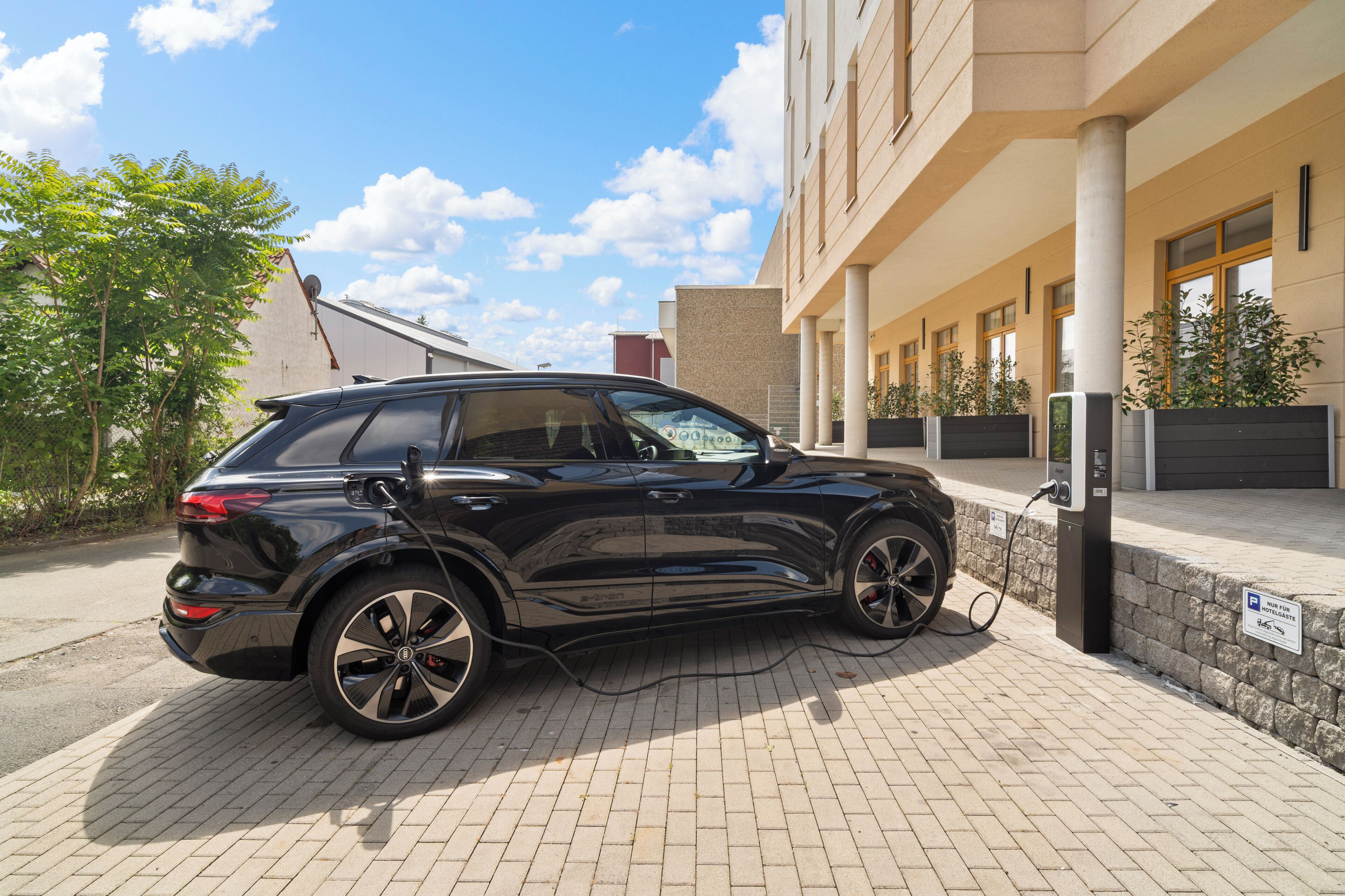 Um SUV preto estacionado em uma estação de carregamento para veículos elétricos, com um edifício e árvores ao fundo.