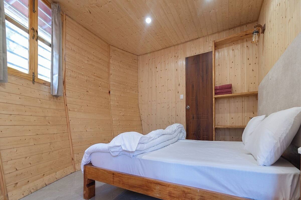 Quarto de madeira com uma cama, uma porta e uma prateleira com toalhas.