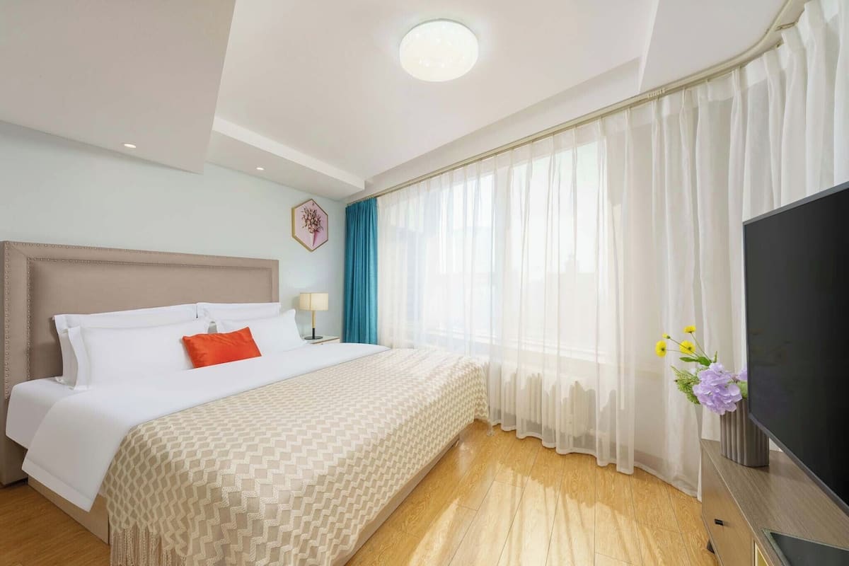 Quarto de hotel com cama, televisão, mesa de cabeceira e janela com cortinas leves.