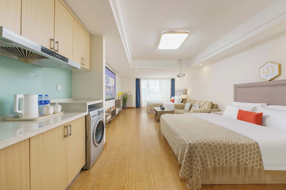 Quarto de hotel com cama, escrivaninha, cadeira, máquina de lavar e cozinha compacta.
