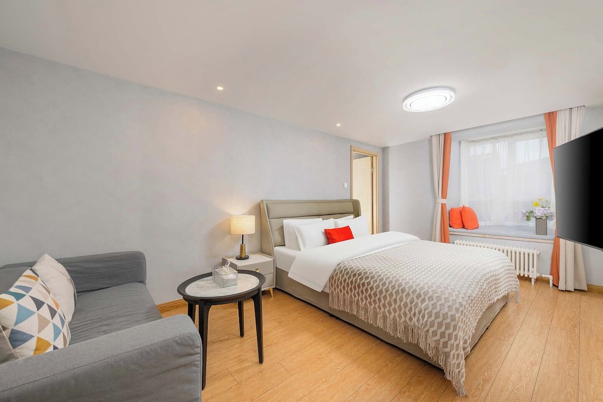 Quarto de hotel com cama, sofá, mesa de cabeceira, luminária, televisão e janela com cortinas.
