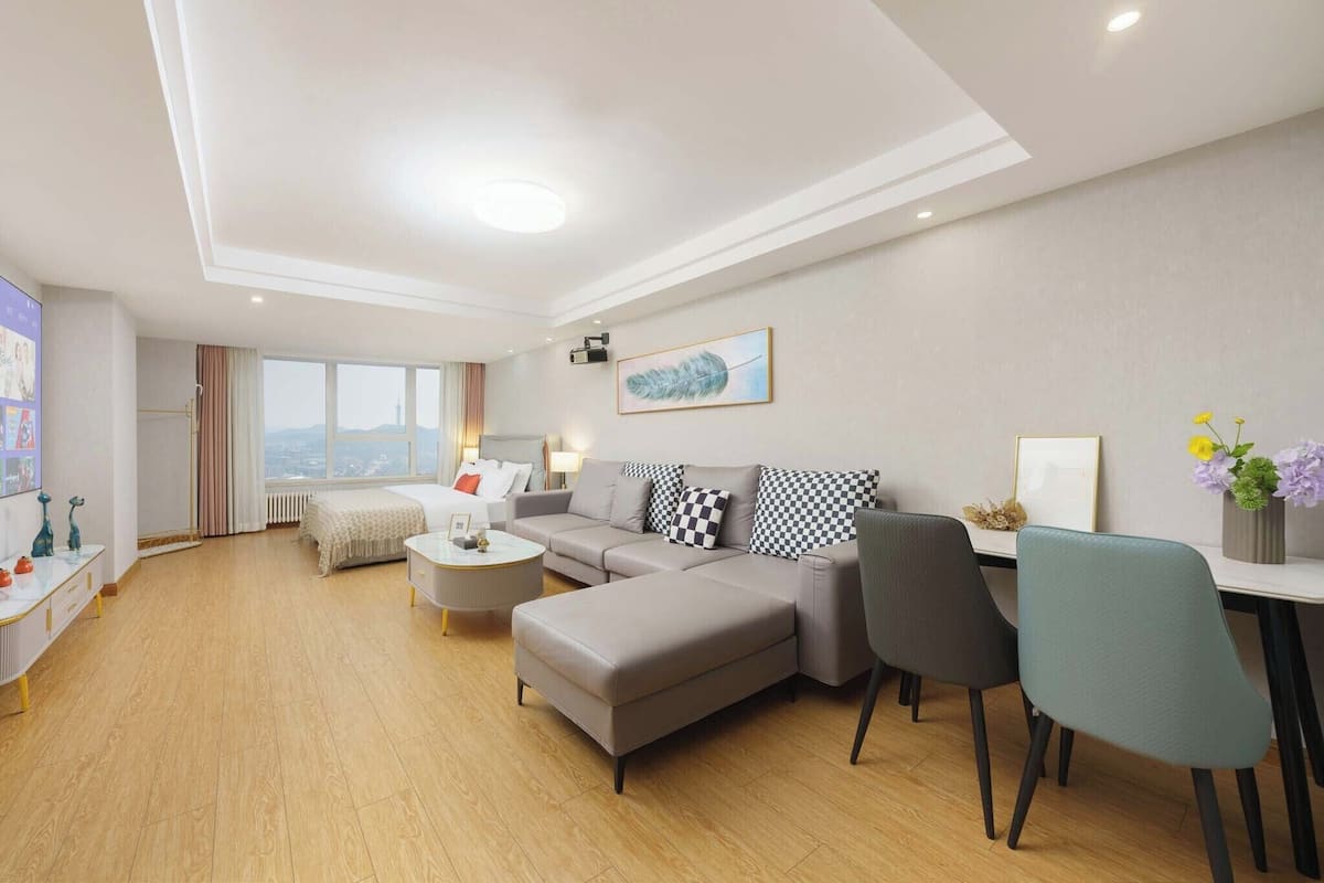 Uma sala de estar moderna com sofá, banquinho e poltronas, uma área de jantar com mesa e uma grande janela com vista.