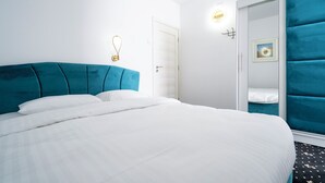 Uma cama com cabeceira azul-turquesa, roupa de cama branca, uma porta branca e um guarda-roupa azul-turquesa com um quadro na parede.