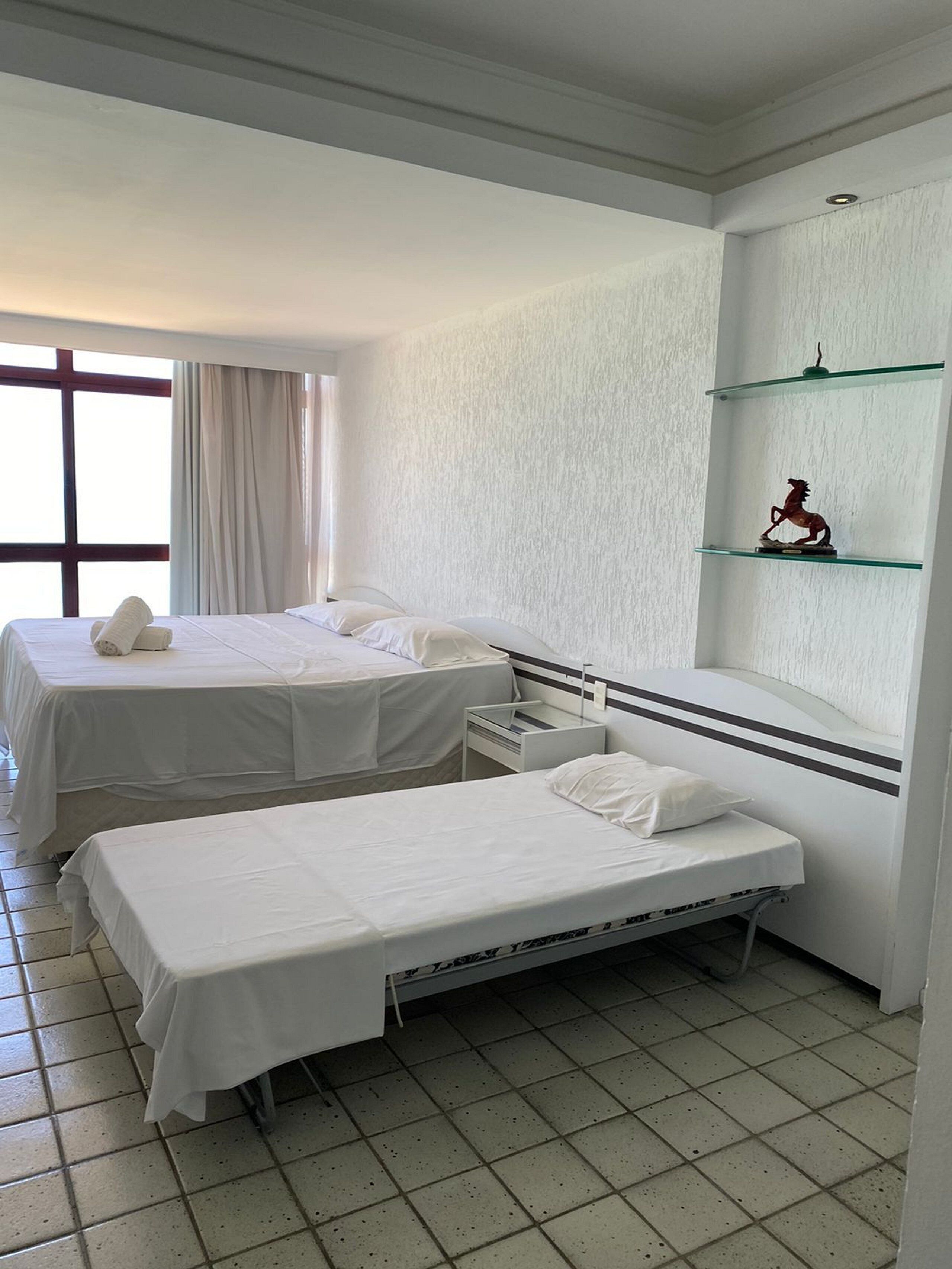 Quarto de hotel com duas camas, uma pequena mesa de cabeceira, prateleira na parede com uma estátua e uma janela grande com cortinas.